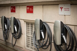 Uma fileira de estações de carregamento para veículos elétricos Tesla fixadas numa parede, com placas a indicar 'Tesla Vehicles' e 'Veículos Elétricos'.