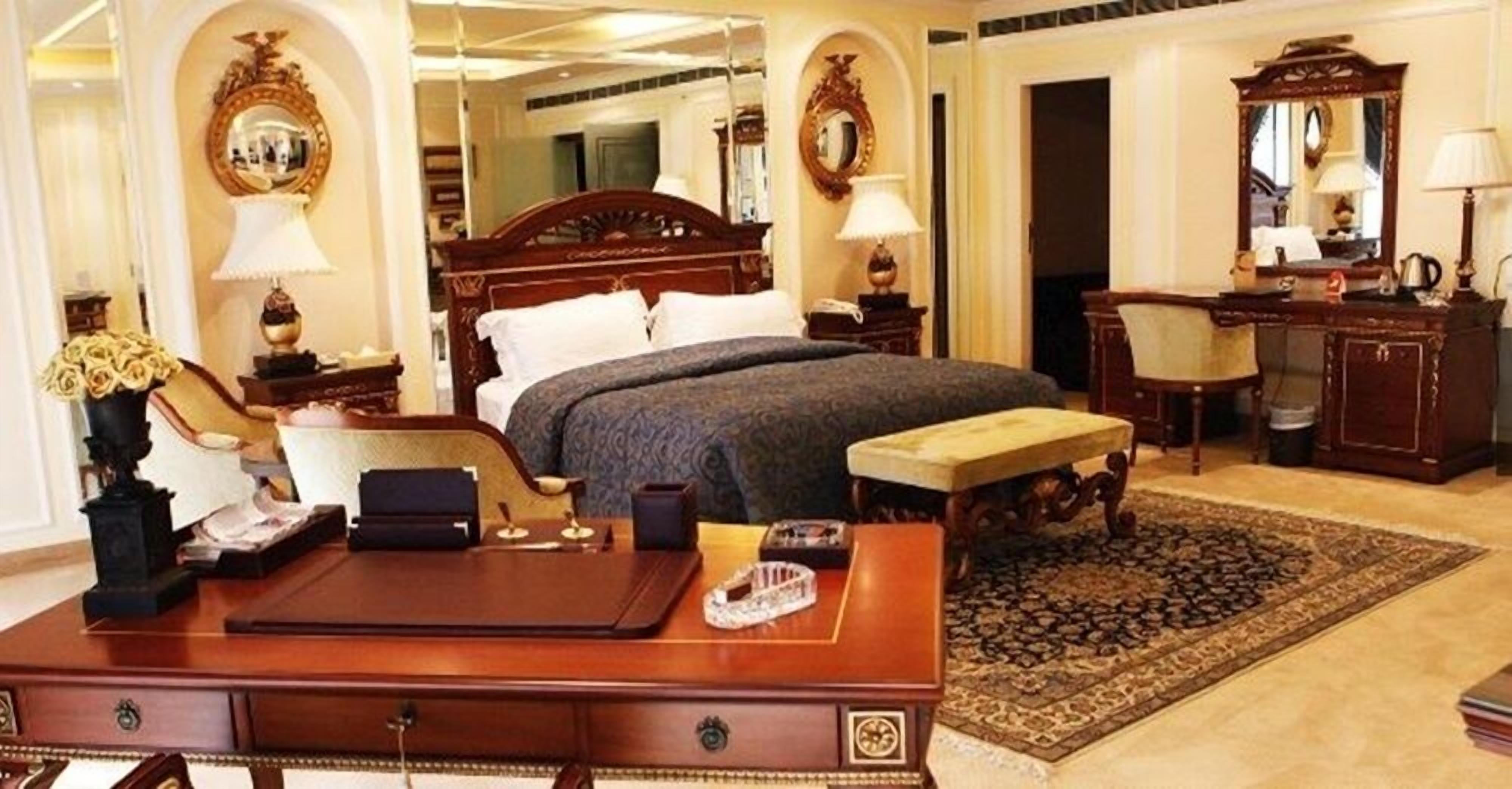 Quarto de hotel com uma cama grande, uma escrivaninha com cadeira, uma toucador com espelho e um tapete grande.
