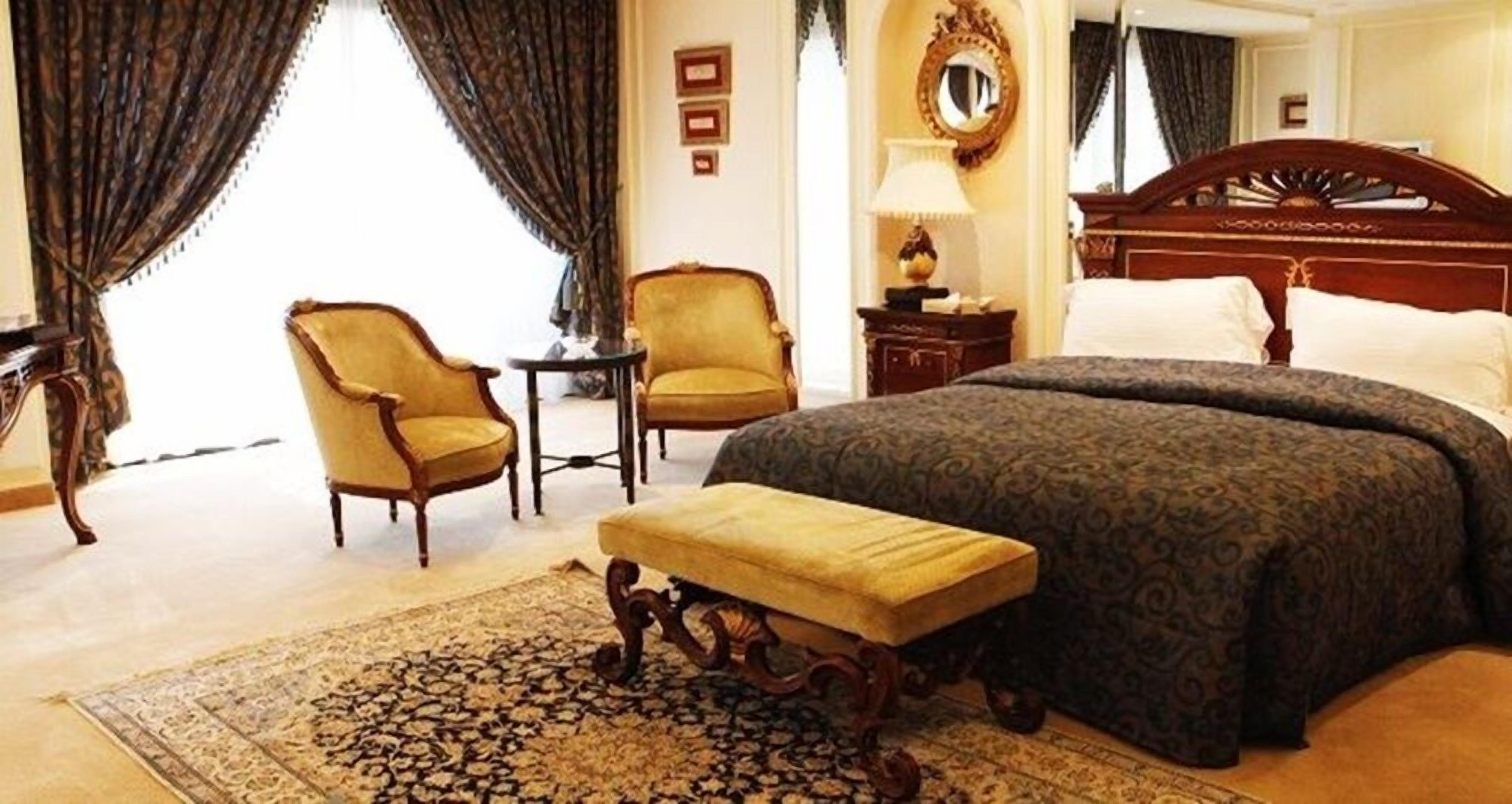 Quarto de hotel com uma cama grande, duas poltronas, uma mesa pequena, um banco e um tapete estampado.