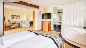 Quarto de hotel moderno com uma cama grande, área de estar com sofá e cadeiras, televisão, chuveiro em box de vidro e piso de madeira.