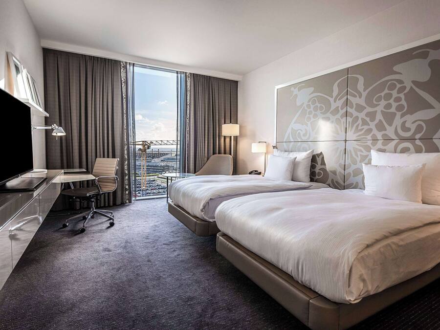 Quarto de hotel com uma cama grande, uma escrivaninha com televisão, uma cadeira e vista para a cidade.