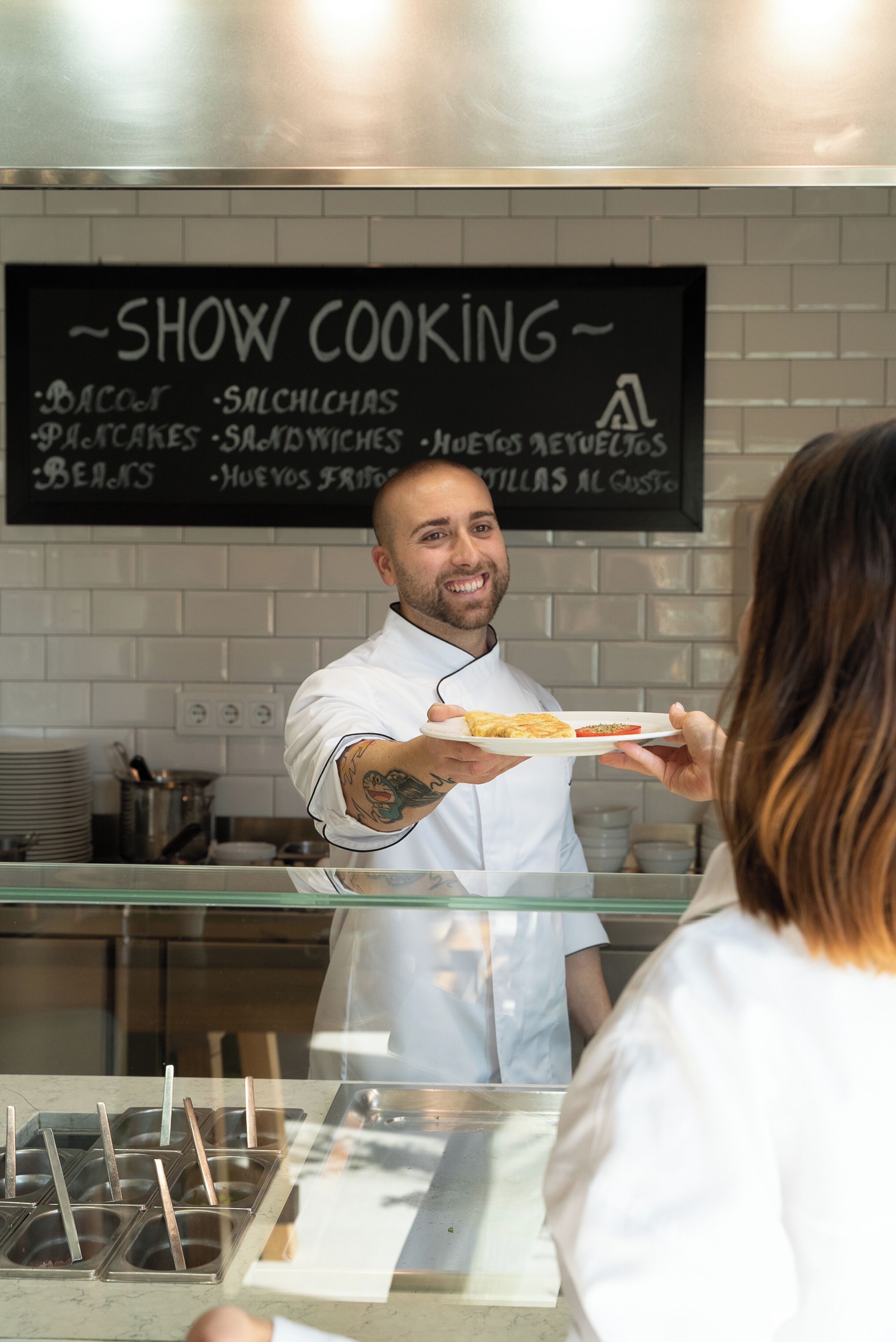 Um chef serve um prato para um cliente em um restaurante, com um quadro de menu ao fundo listando itens como bacon, panquecas e sanduíches.