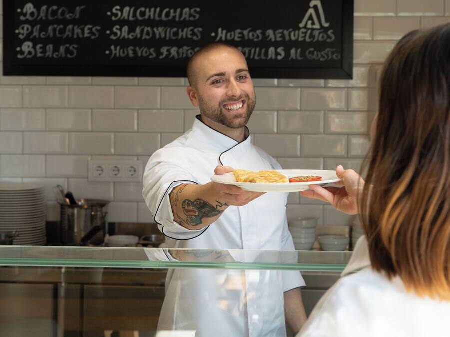 Um chef serve um prato para um cliente em um restaurante, com um quadro de menu ao fundo listando itens como bacon, panquecas e sanduíches.
