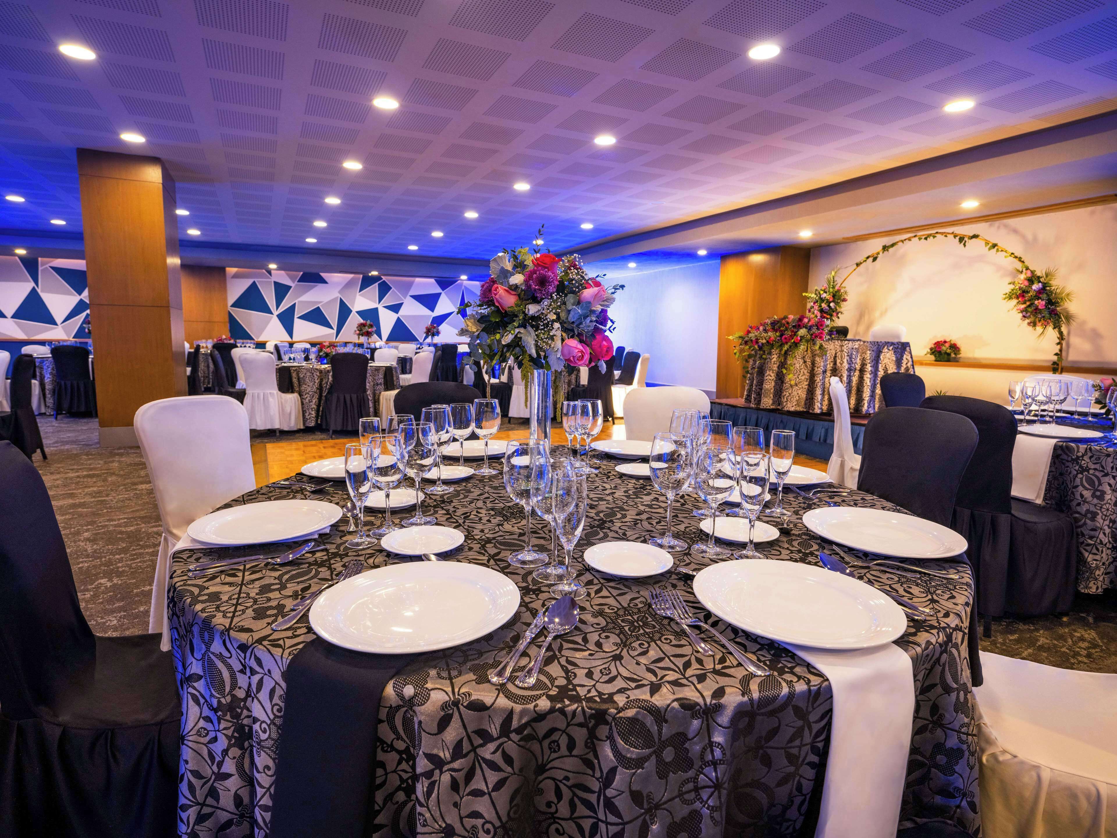 Uma sala de banquetes com mesas redondas preparadas para um evento formal, apresentando um design geométrico no teto e arranjos florais.