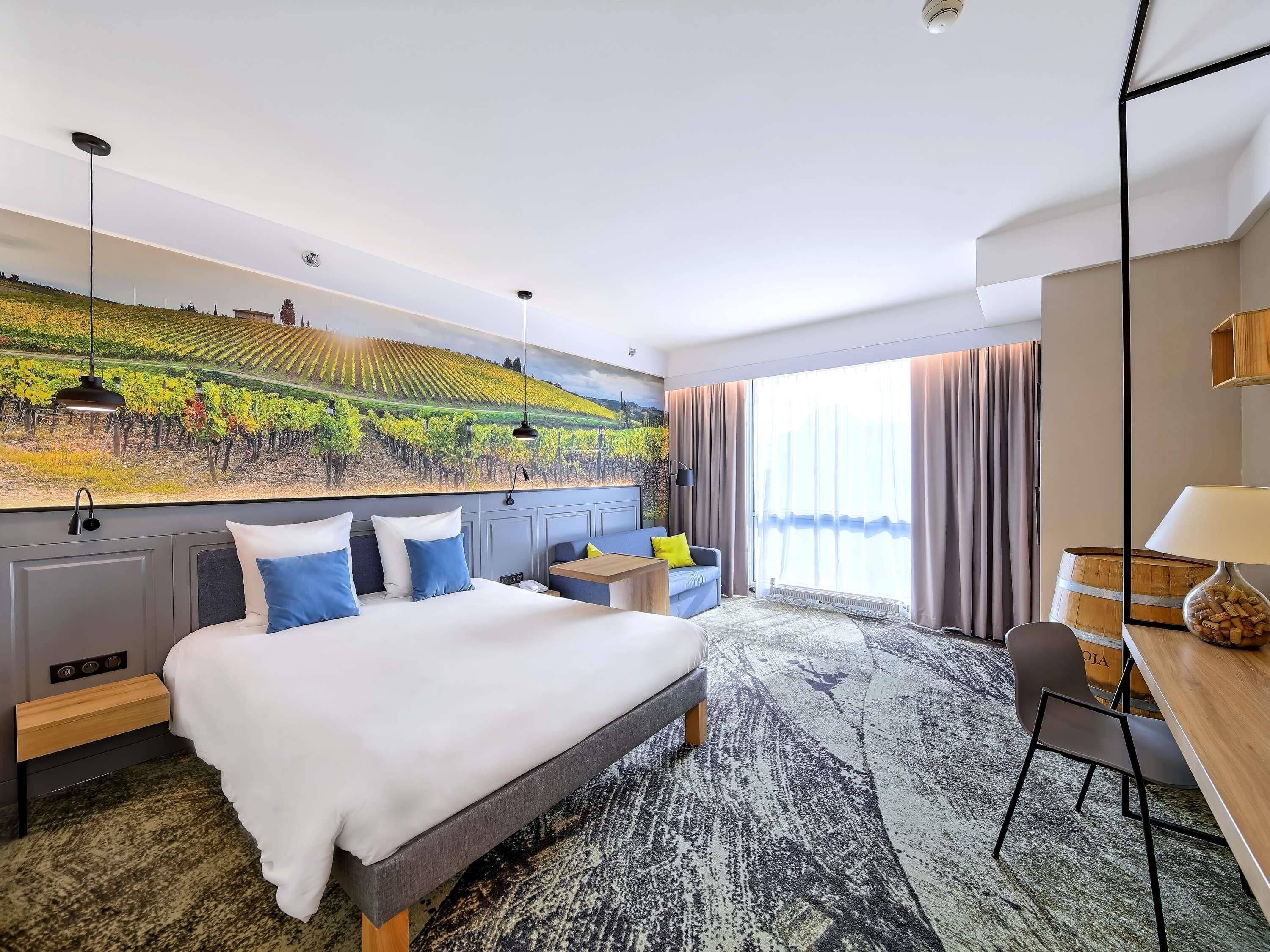 Quarto de hotel com cama, escrivaninha, cadeira, janela grande e um mural na parede.
