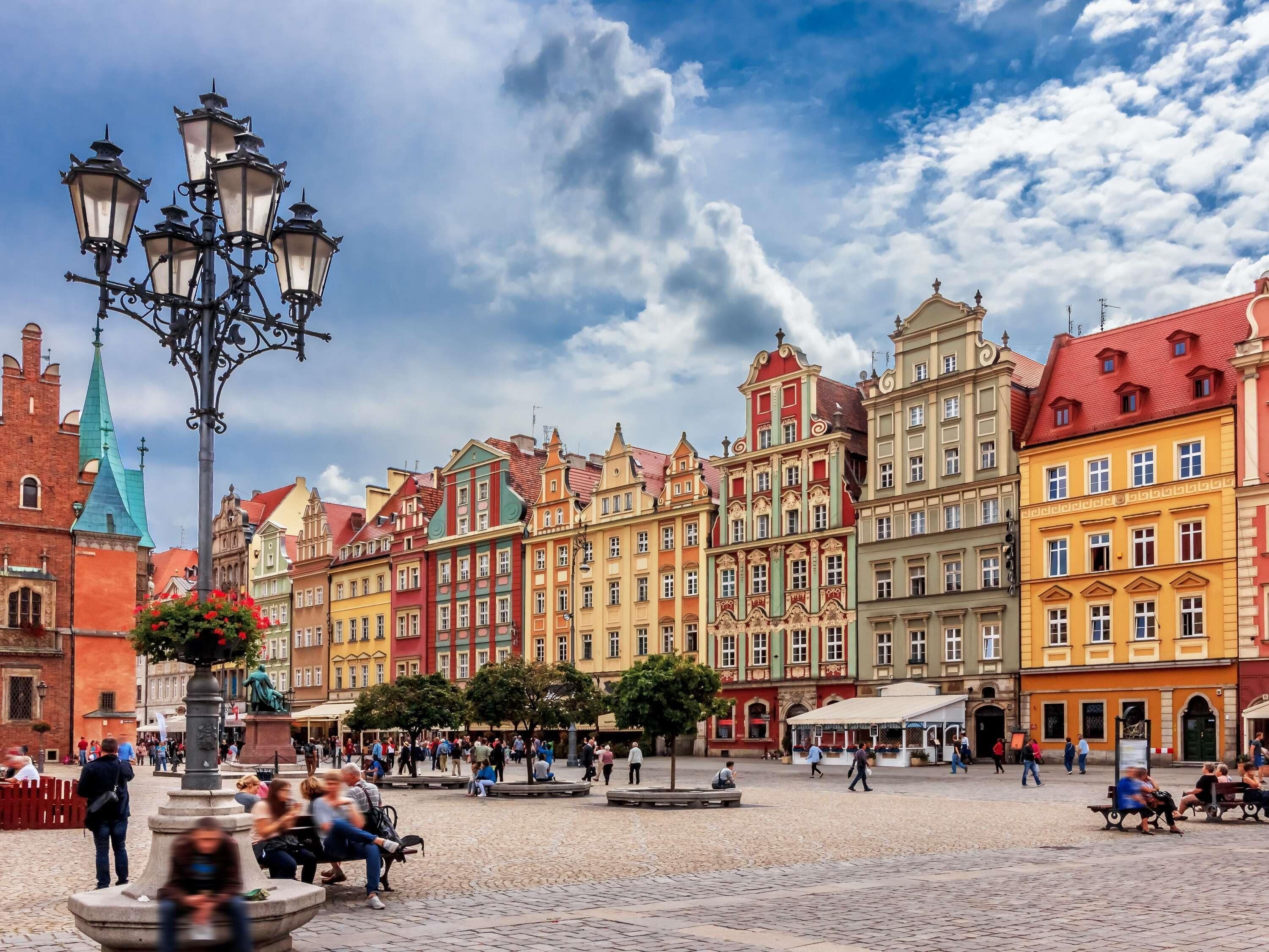 Uma praça histórica com calçamento de pedra, uma fonte central e edifícios coloridos com arquitetura tradicional.