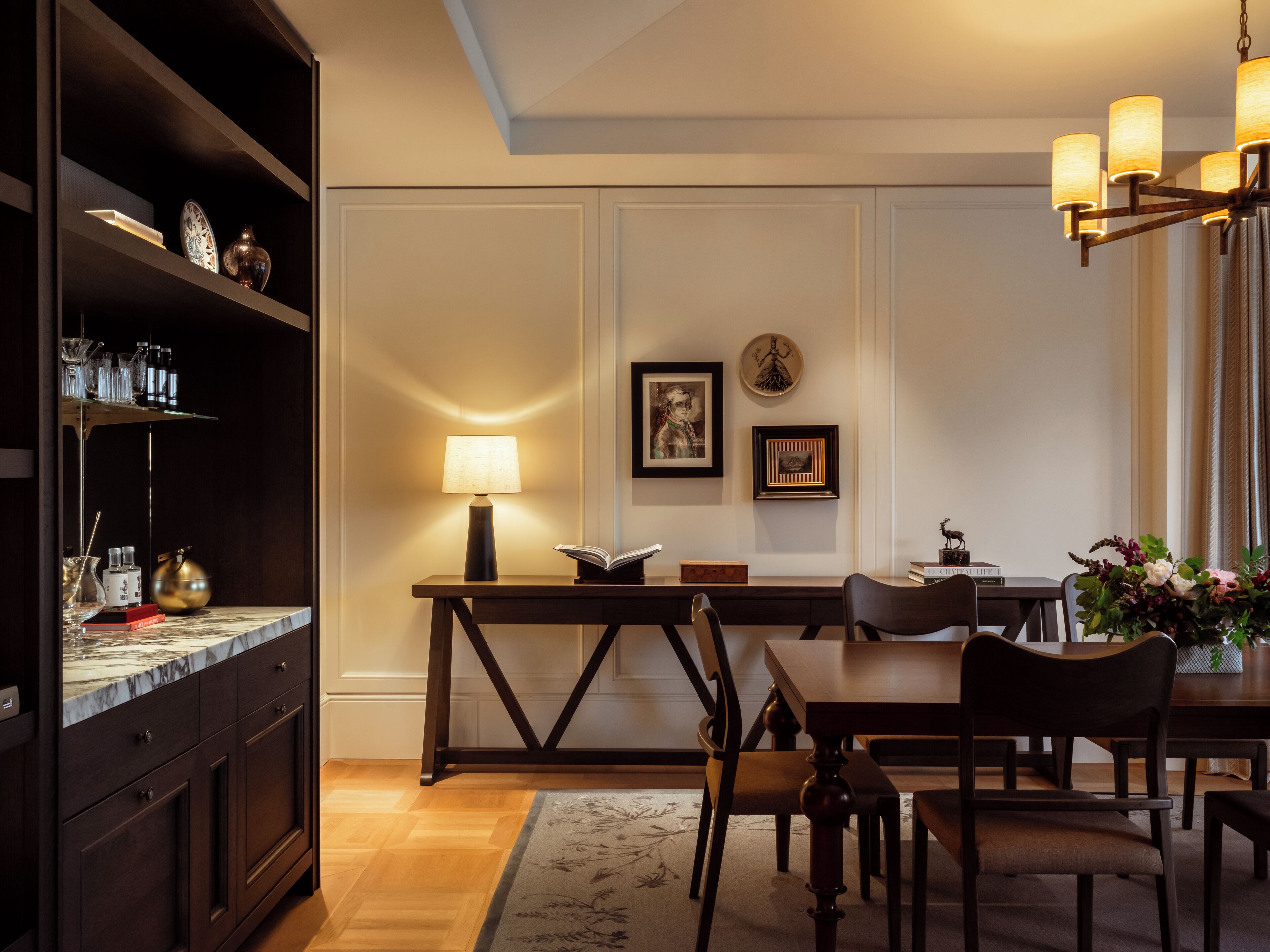 Uma sala de jantar com mesa de madeira, cadeiras, um lustre e uma estante com objetos decorativos.