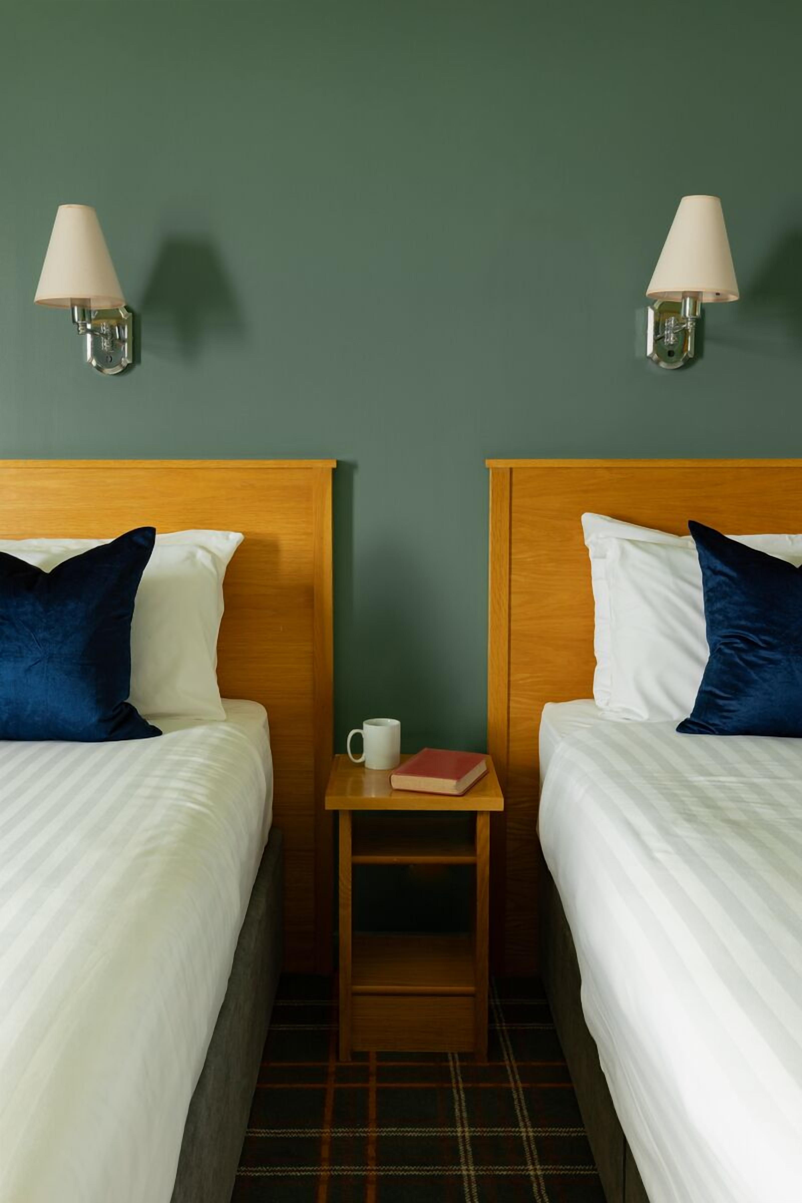 Quarto de hotel com duas camas, cada uma com cabeceira de madeira, roupa de cama branca e almofadas azuis. Há duas luminárias fixadas na parede e uma mesa de cabeceira com um livro e uma caneca.
