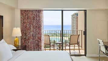 Quarto de hotel com uma cama, uma mesa de cabeceira com abajur, uma varanda com vista para o oceano e edifícios, e duas cadeiras.