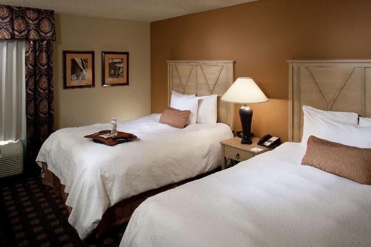 Quarto de hotel com duas camas, uma mesa de cabeceira com abajur, um telefone e duas fotos emolduradas na parede.