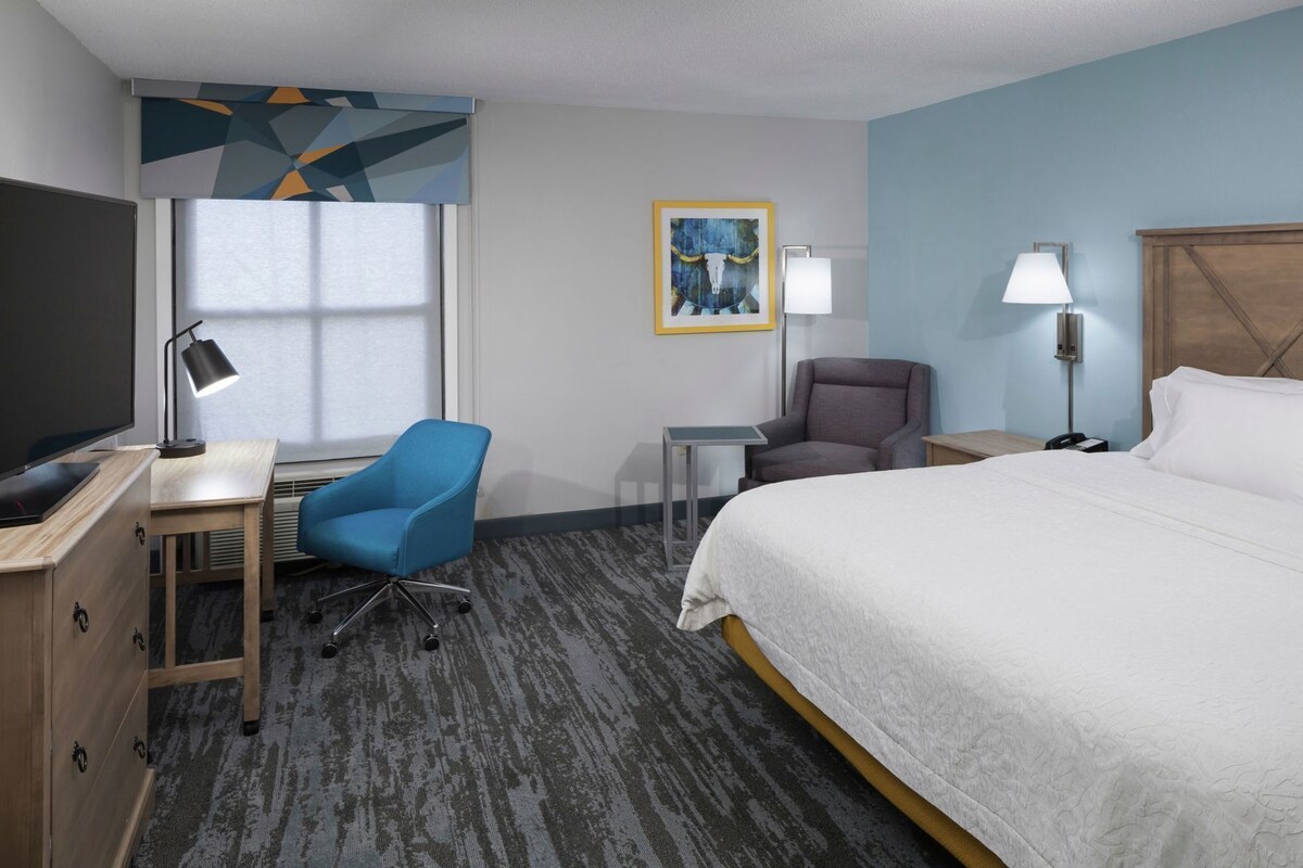 Quarto de hotel com cama, escrivaninha, cadeira, televisão, luminárias e obras de arte.
