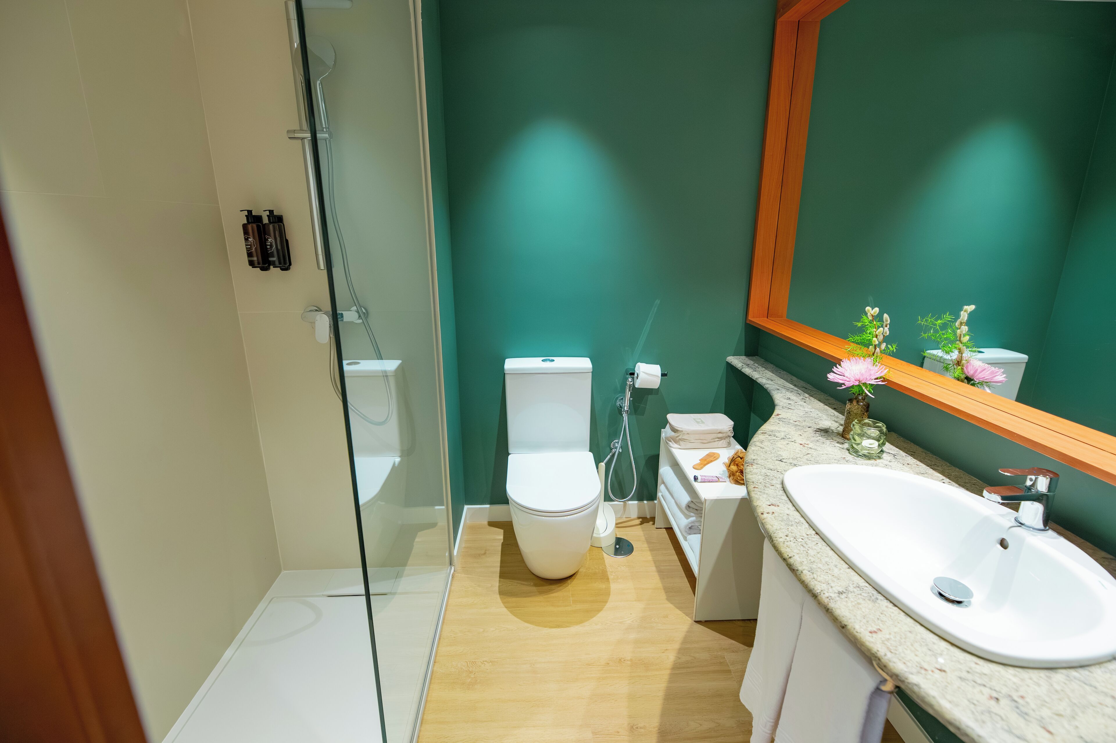 Um banheiro com box de vidro, vaso sanitário branco, pia com bancada de mármore e espelho com moldura de madeira.