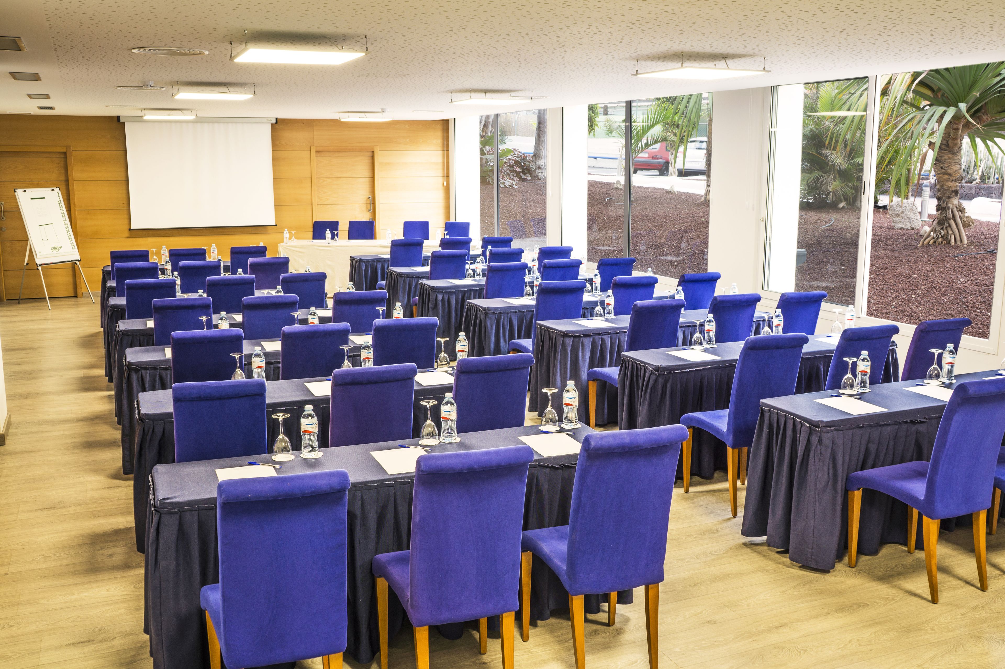 Uma sala de conferências com fileiras de mesas e cadeiras, um quadro branco e janelas amplas com vista para palmeiras e um estacionamento.