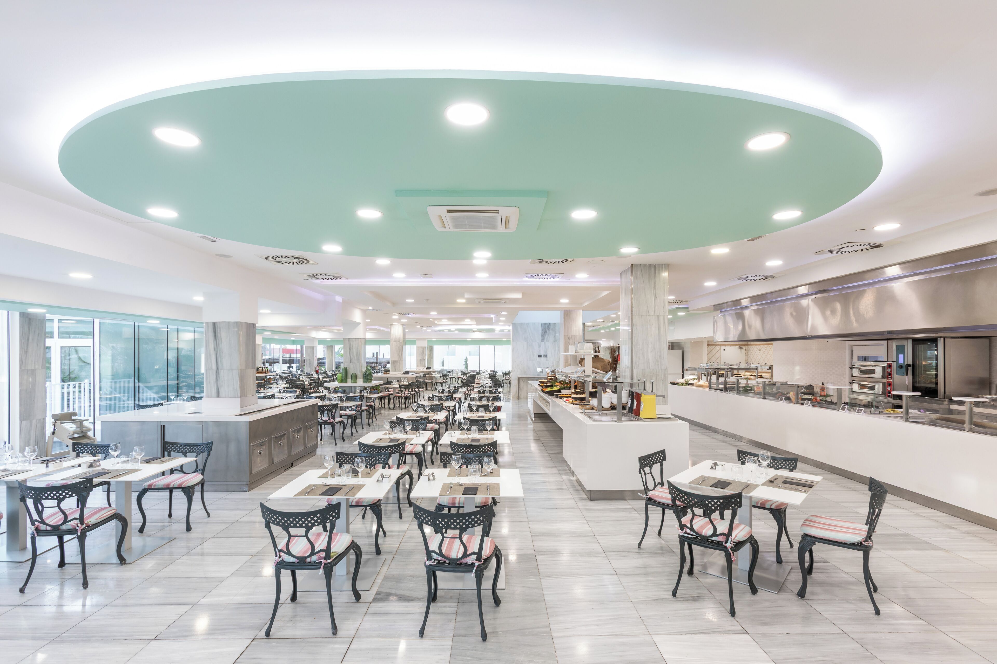 Uma ampla sala de jantar interna com várias mesas e cadeiras, um balcão de serviço central e uma área de cozinha ao fundo.