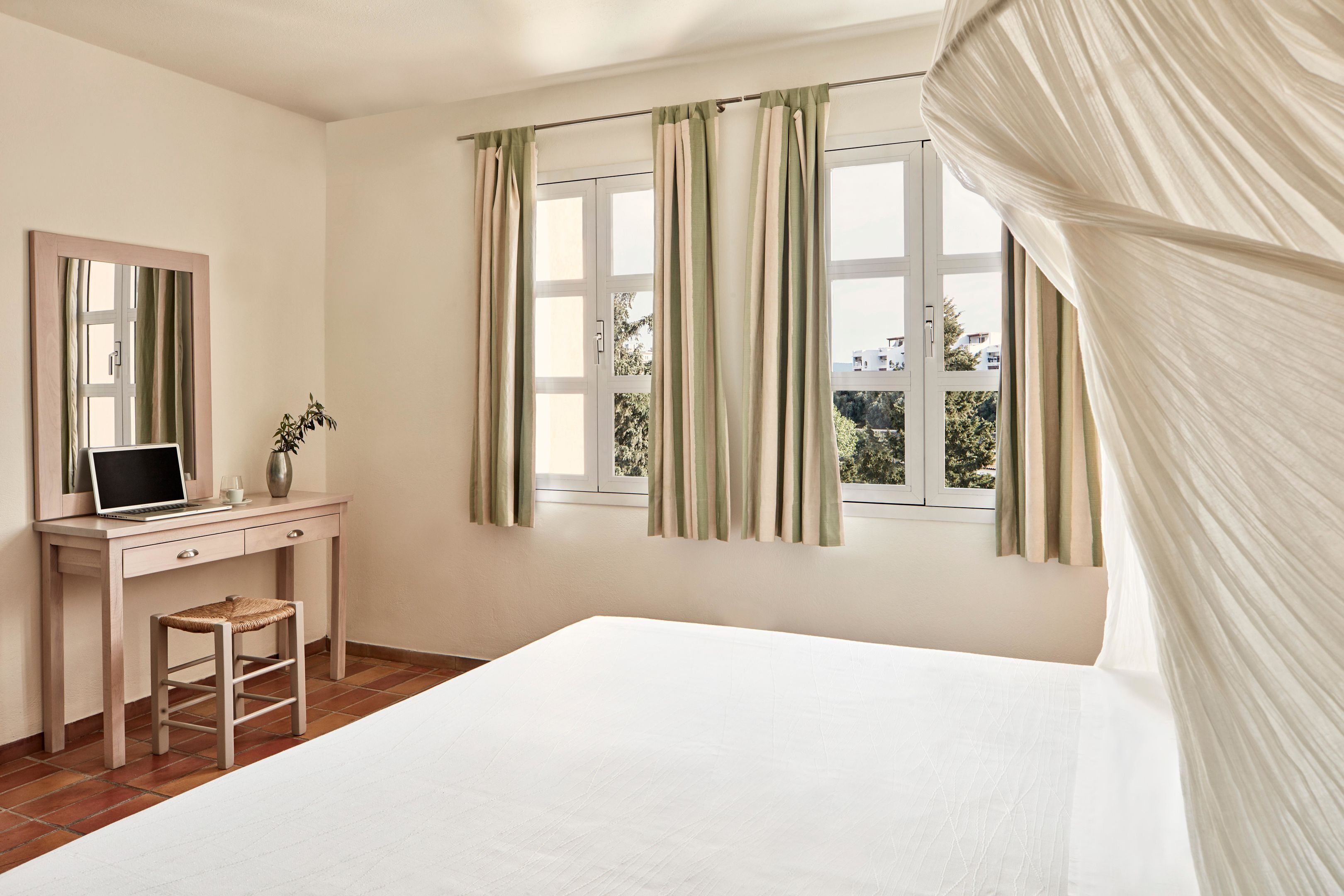 Um quarto com cama, escrivaninha com laptop, cadeira e janela com cortinas.