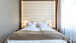 Cama bem arrumada, com esquema de cores bege e branco, cabeceira de madeira e luminárias laterais.