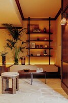 Quarto com um banco de madeira, uma mesa redonda e uma estante com objetos decorativos.