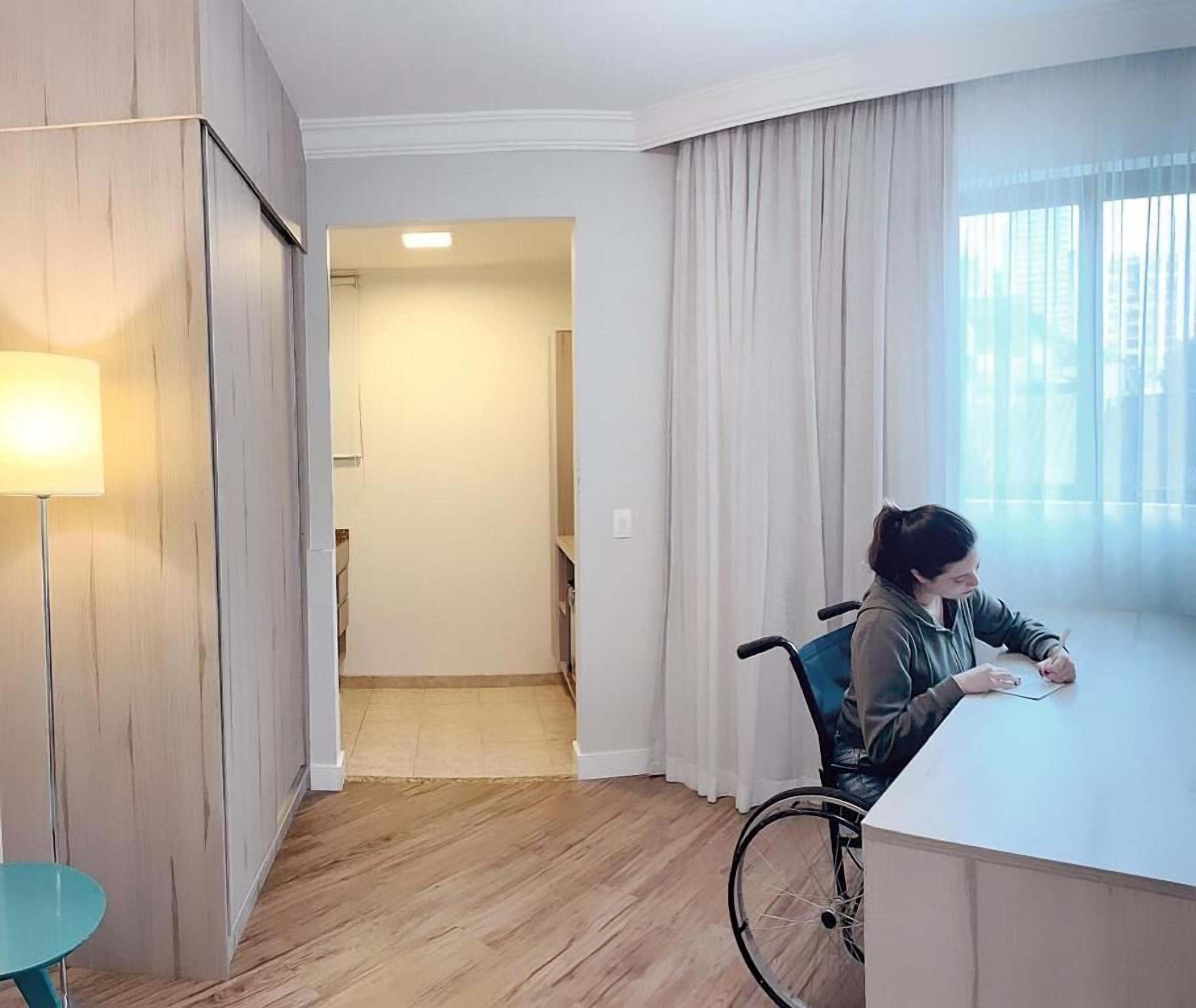Quarto acessível em cadeira de rodas, com escrivaninha, luminária e vista da cidade através de amplas janelas.