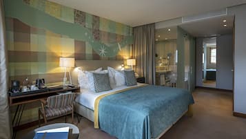 Quarto de hotel com cama, luminárias de cabeceira, escrivaninha com cadeira, espelho e banheiro visível por uma porta aberta.