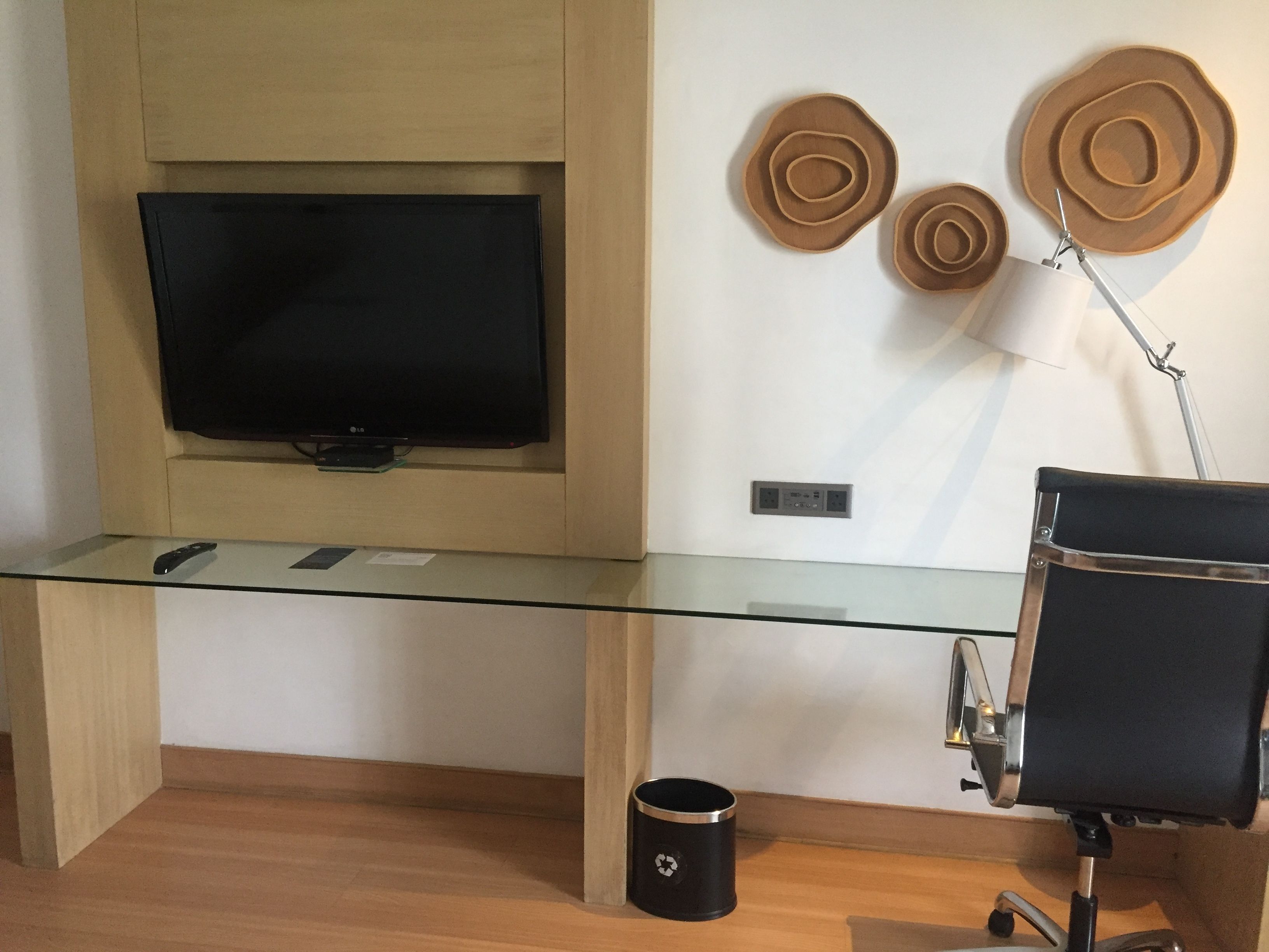 Sala moderna com televisão de tela plana presa a uma parede de madeira, mesa de centro de vidro, cadeira de escritório preta e duas placas decorativas penduradas na parede.