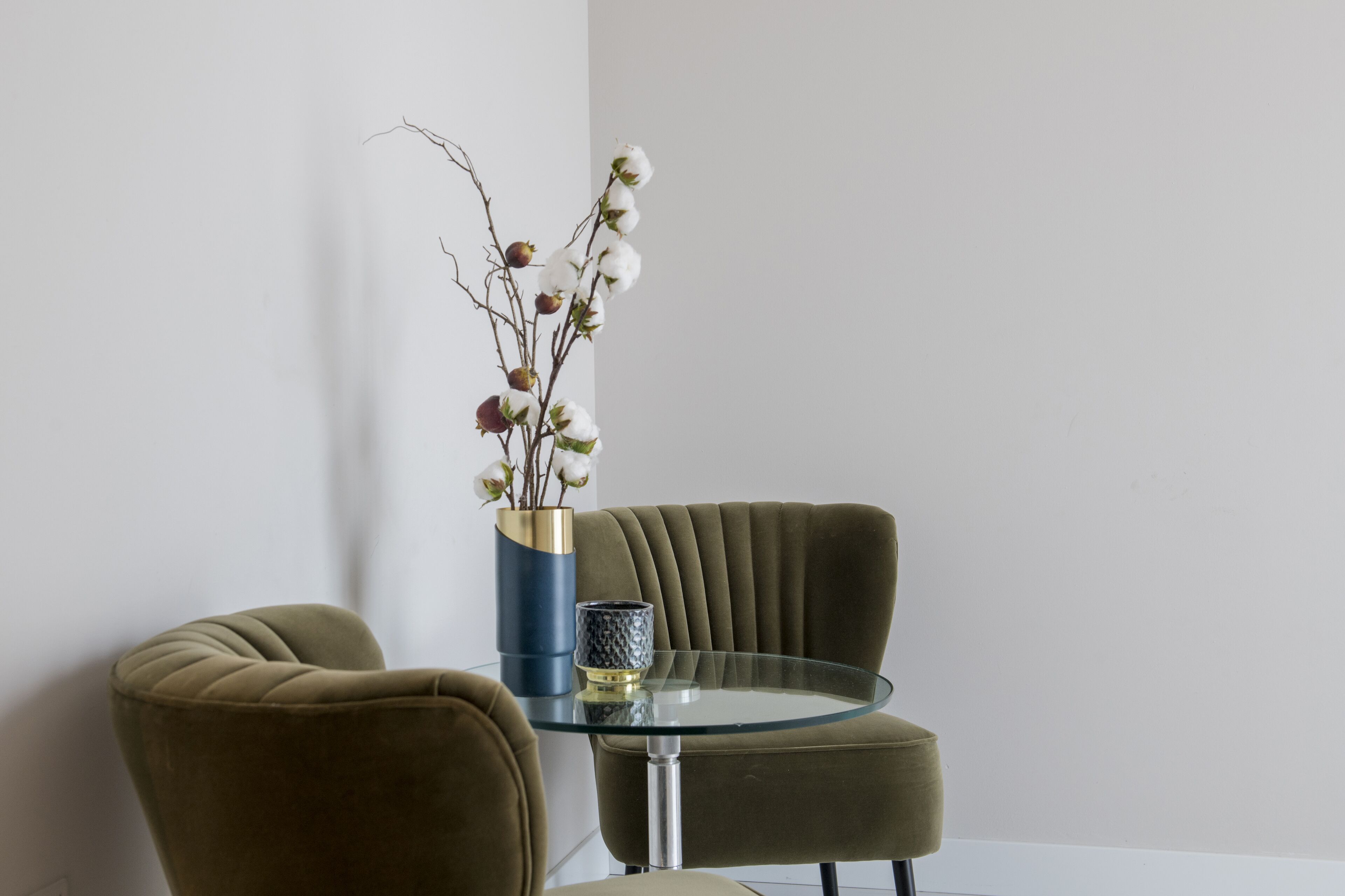 Quarto moderno com mesa de vidro redonda, duas cadeiras estofadas, um vaso com galhos e algodão, e um recipiente texturizado.
