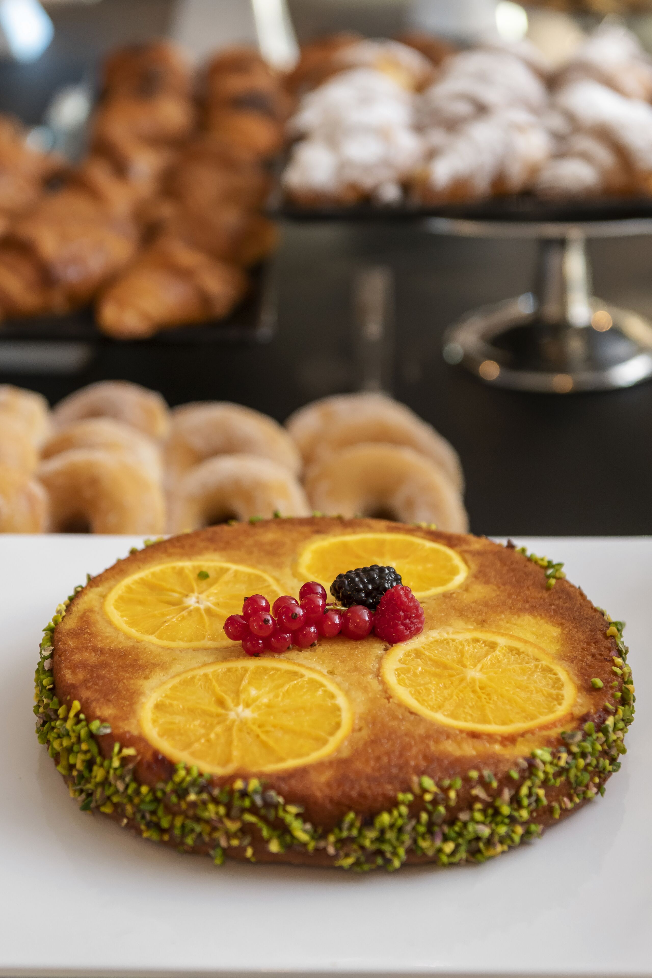 Um bolo redondo com fatias de laranja e frutas vermelhas por cima, rodeado por outros doces.
