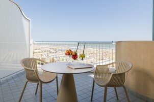 Varanda com duas cadeiras e uma mesa, com vista para uma praia com guarda-sóis e céu claro.