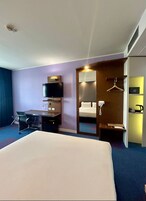 Quarto de hotel com cama, escrivaninha, cadeira, televisão, espelho e guarda-roupa.