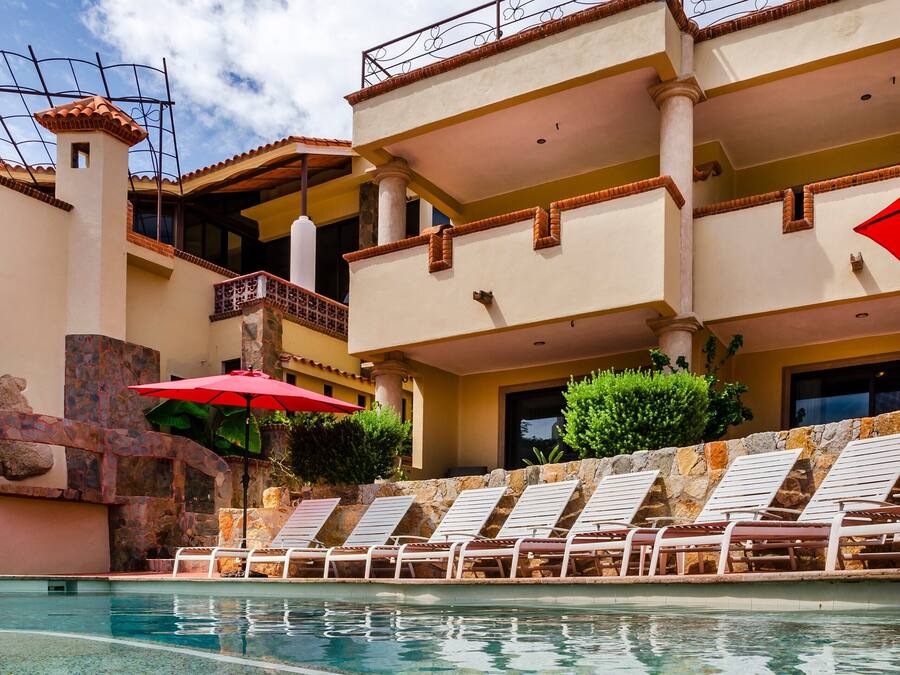 Uma área de piscina com espreguiçadeiras e guarda-sóis, um muro de pedra e um edifício com várias varandas.