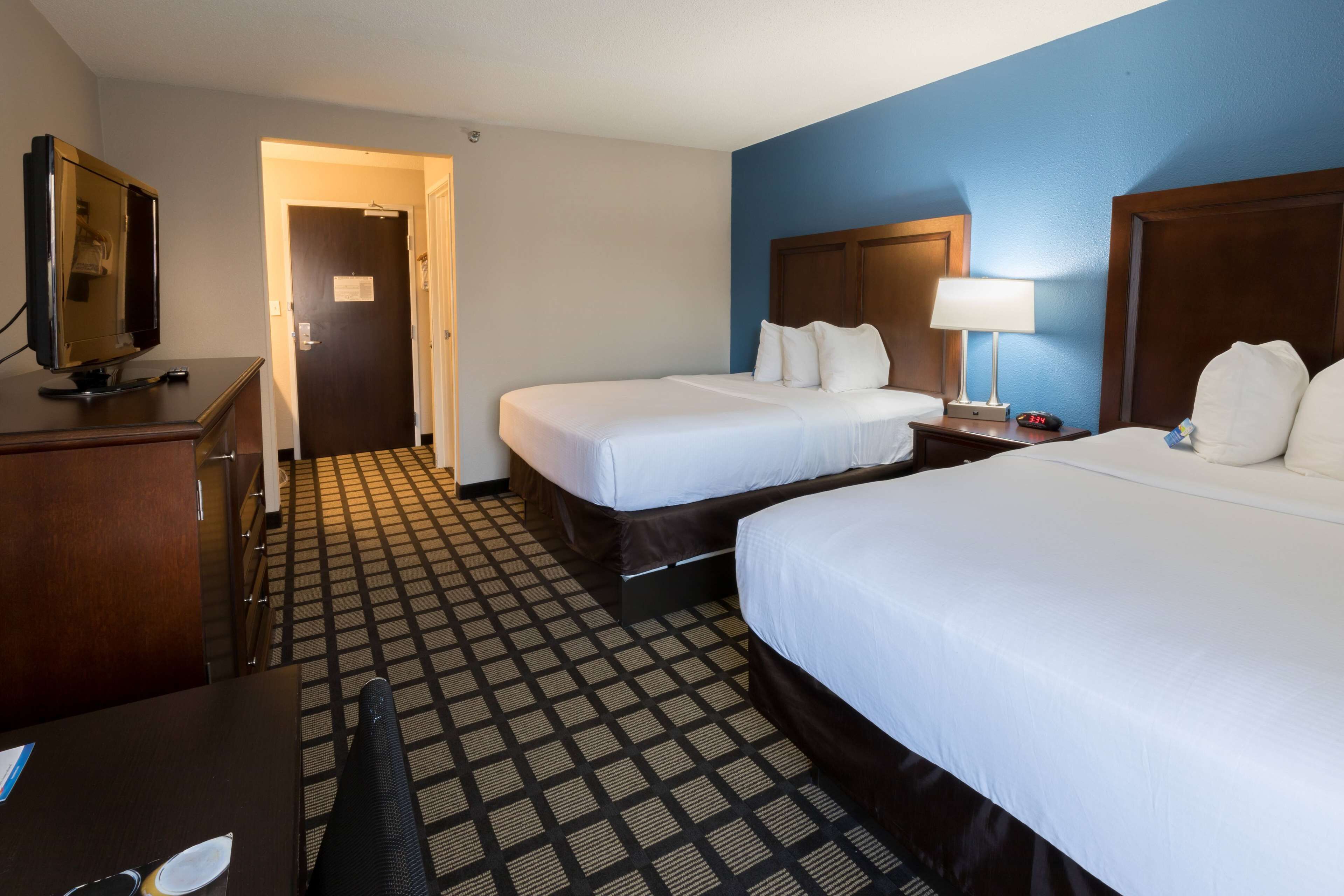 Quarto de hotel com duas camas, uma escrivaninha com cadeira, televisão e luminária.