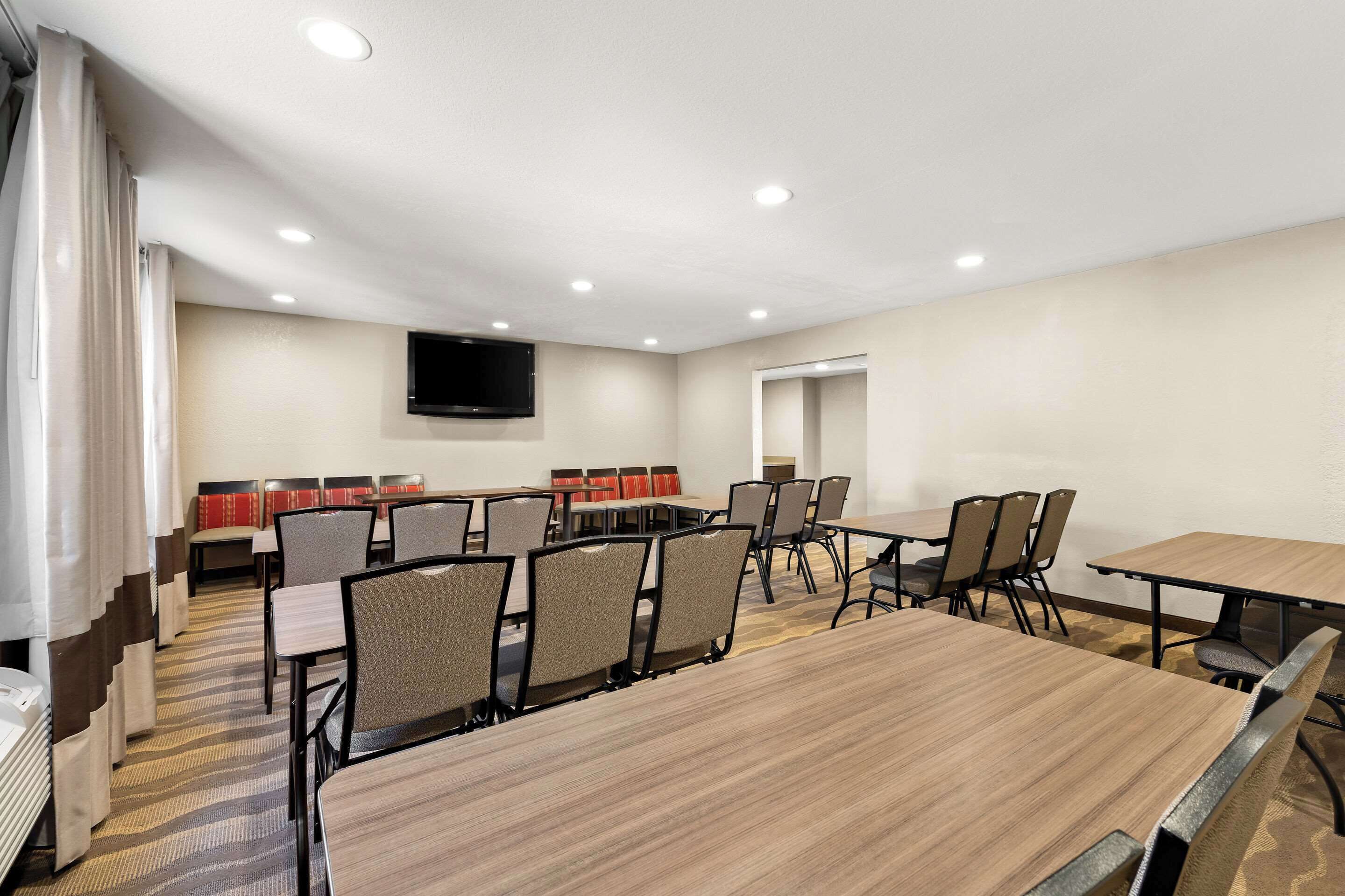 Uma sala de conferências com várias mesas e cadeiras, uma TV de tela plana fixada na parede e piso coberto por carpete.