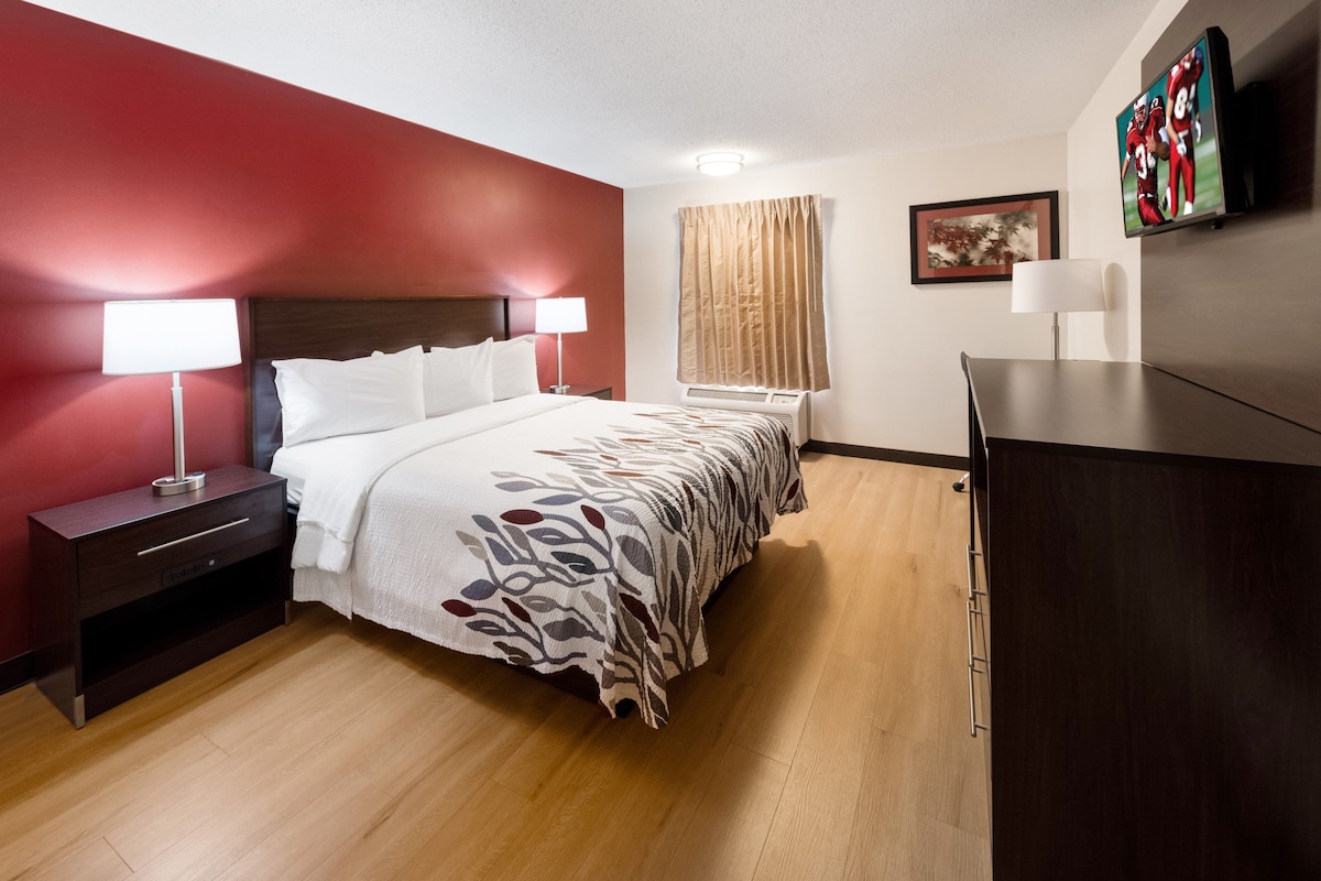 Quarto de hotel com cama, mesas de cabeceira, escrivaninha, cadeira, luminária, janela com cortinas e televisão na parede.