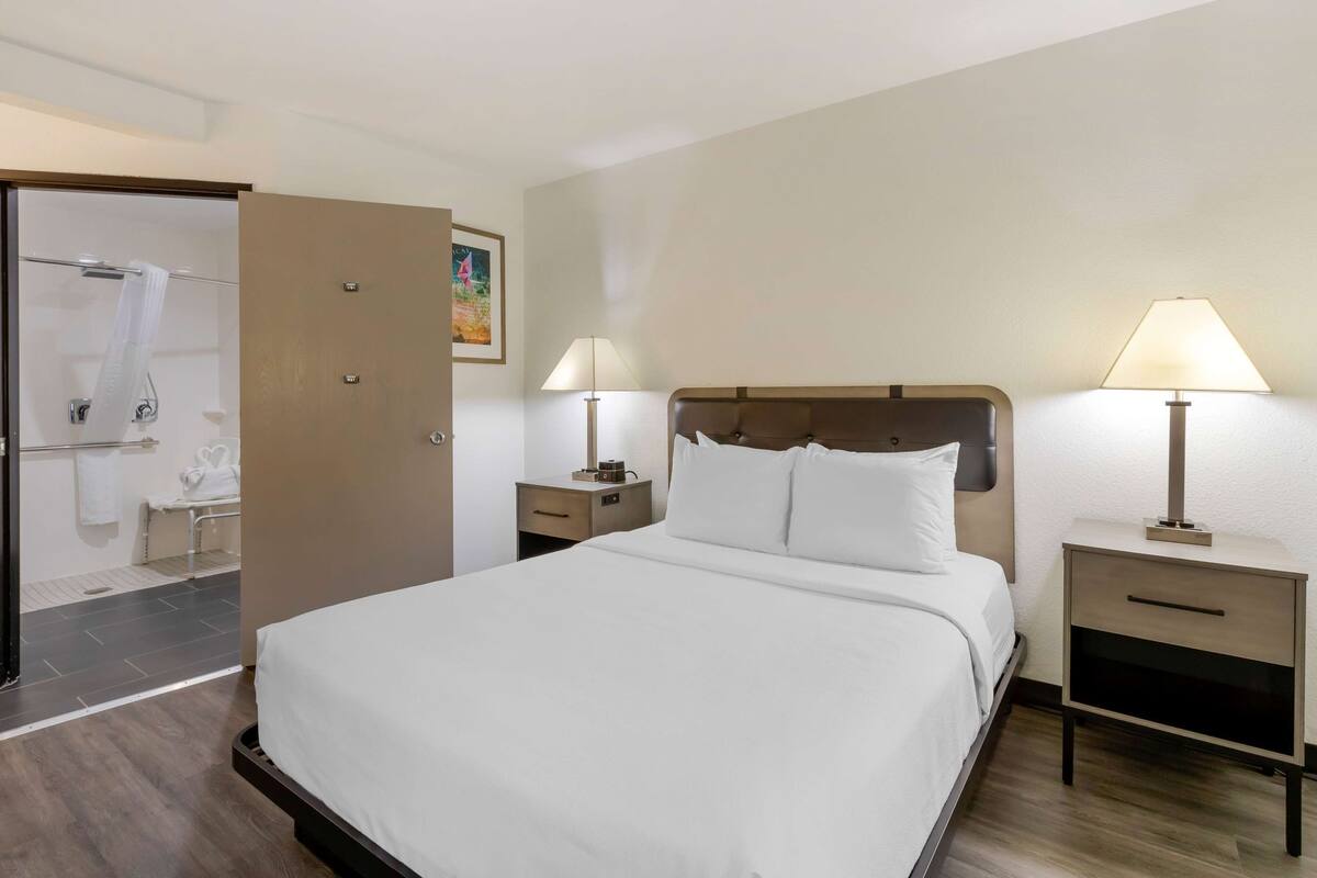Quarto de hotel com uma cama grande, duas mesas de cabeceira com abajures, um banheiro visível através de uma porta aberta e uma pequena mesa com uma cadeira.