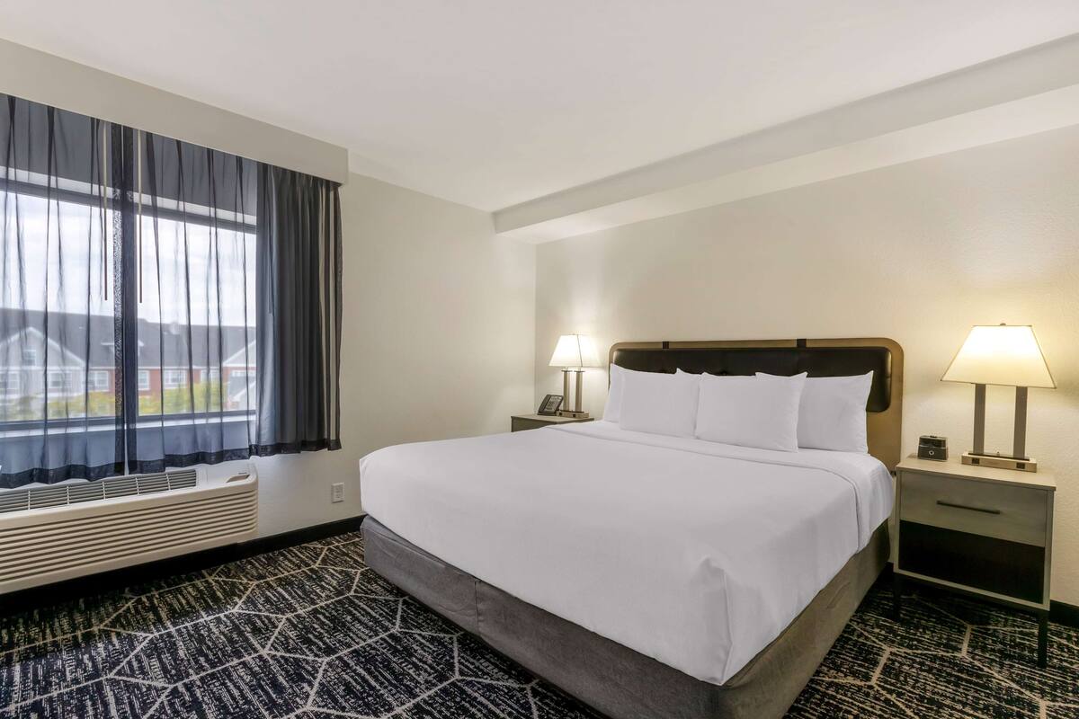 Quarto de hotel com uma cama grande, duas mesas de cabeceira com luminárias, uma janela com cortinas e um tapete estampado.