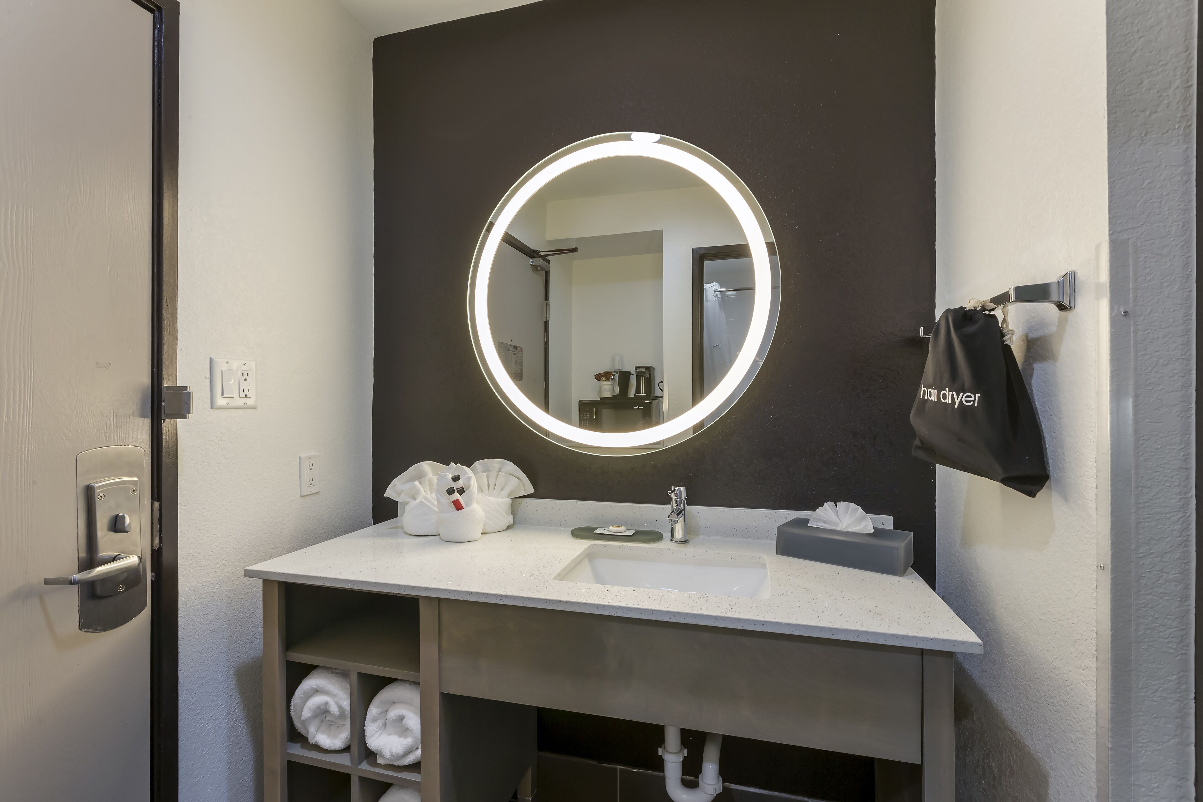 Casa de banho moderna com espelho redondo, lavatório branco, porta-toalhas e uma mala com a etiqueta "secador de cabelo".