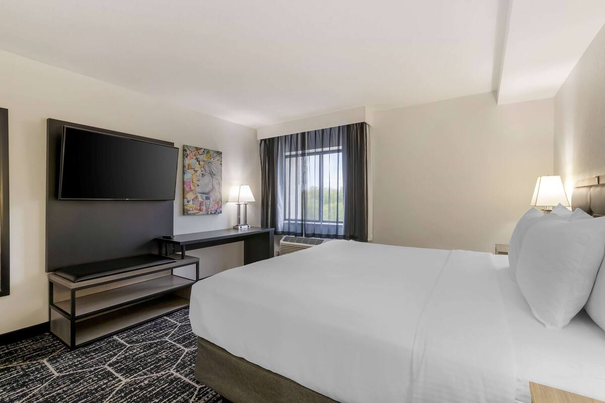Quarto de hotel com uma cama grande, televisão de tela plana na parede, escrivaninha com abajur e janela com cortinas.