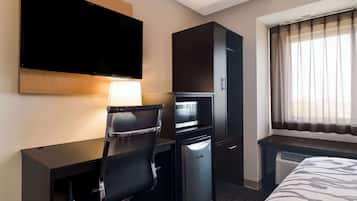 Quarto de hotel com cama, escrivaninha com cadeira, televisão de tela plana na parede, mini-geladeira, micro-ondas e janela com cortinas.