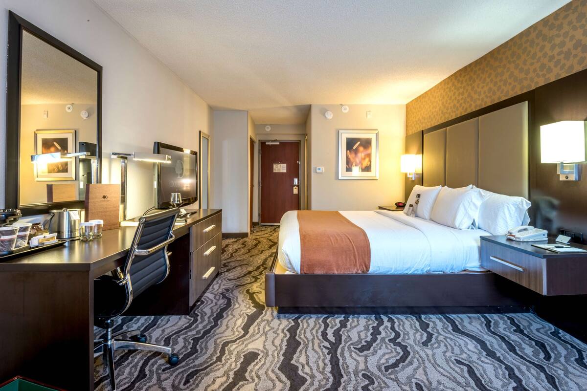 Quarto de hotel com cama, escrivaninha, cadeira e um espelho grande.
