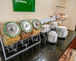 Buffet de café da manhã com granola, barras de cereais, pratos de papel e um espaço para café.