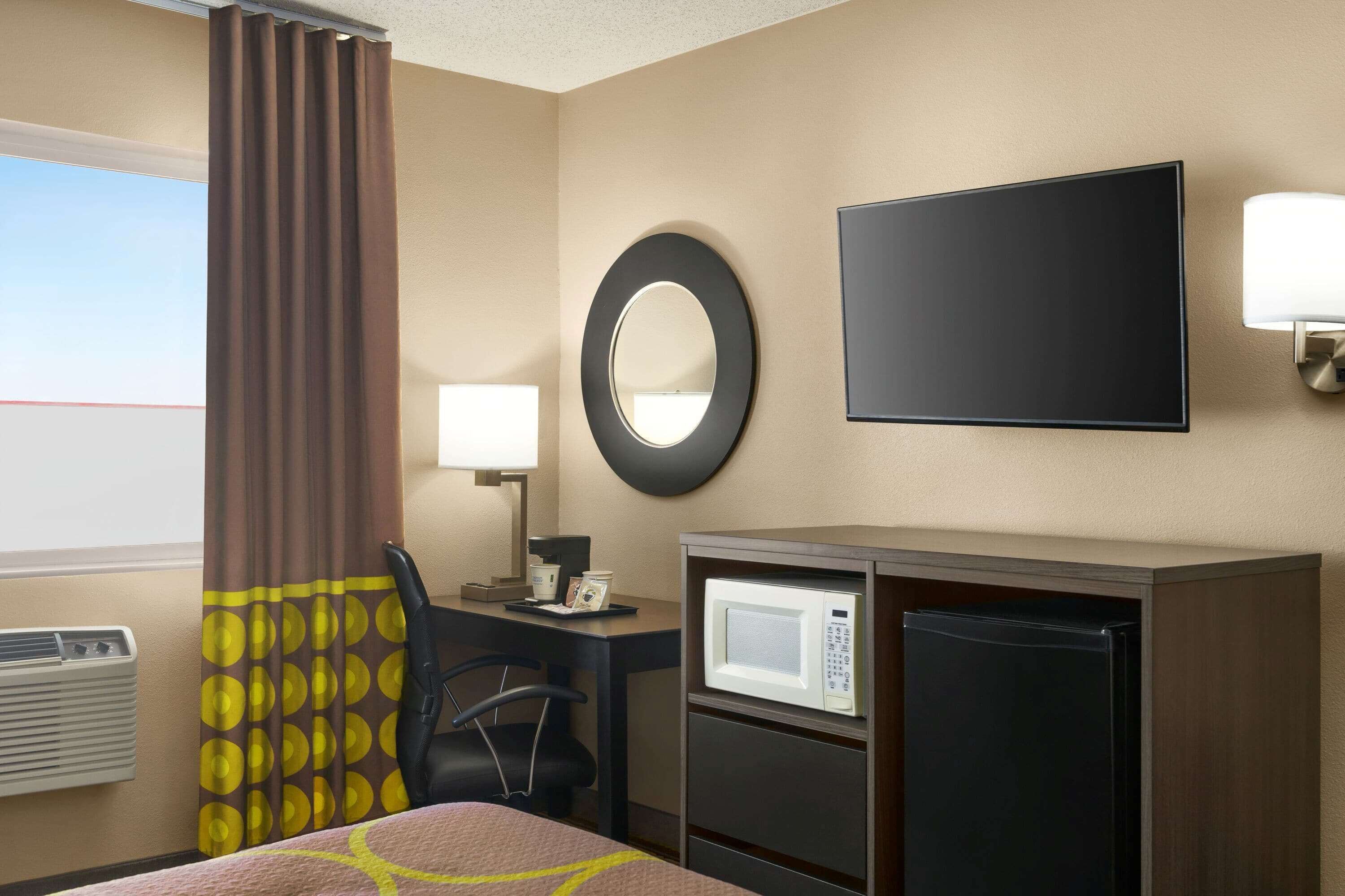 Quarto de hotel com cama, escrivaninha com luminária, micro-ondas, geladeira, espelho e televisão.