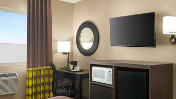 Quarto de hotel com cama, escrivaninha com luminária, micro-ondas, geladeira, espelho e televisão.