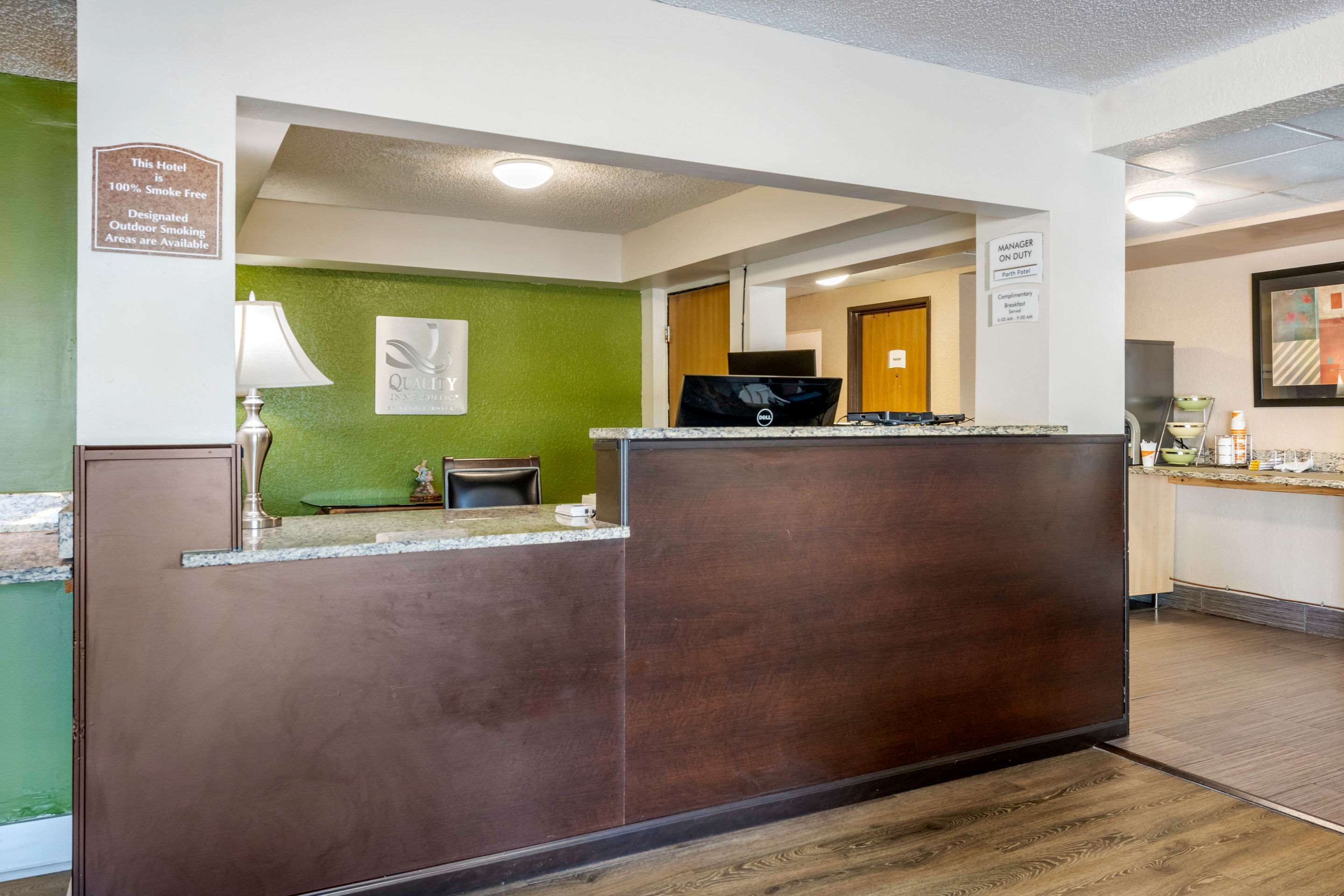 Uma área de recepção de hotel com um balcão de cor castanho escuro, uma parede verde e uma placa indicando ambiente 100% livre de fumo.