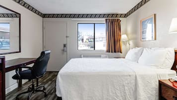 Quarto de hotel com cama, escrivaninha, cadeira, janela com cortinas e luminária na parede.