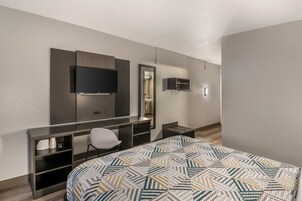 Quarto de hotel com cama, escrivaninha, cadeira, televisão e espelho.
