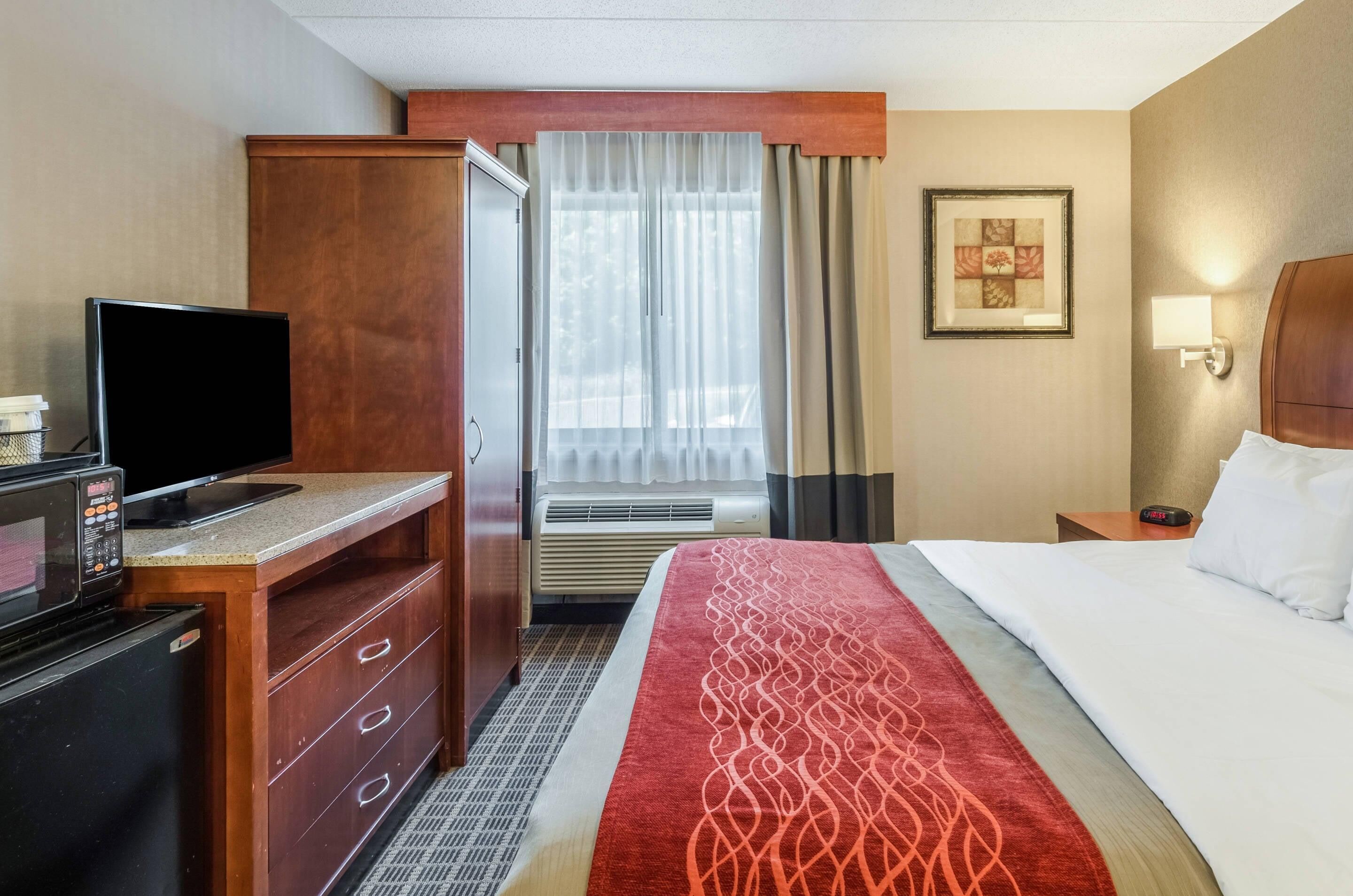 Quarto de hotel com cama, escrivaninha com televisão, minibar, guarda-roupa, ar condicionado, janela com cortinas e quadro pendurado na parede.