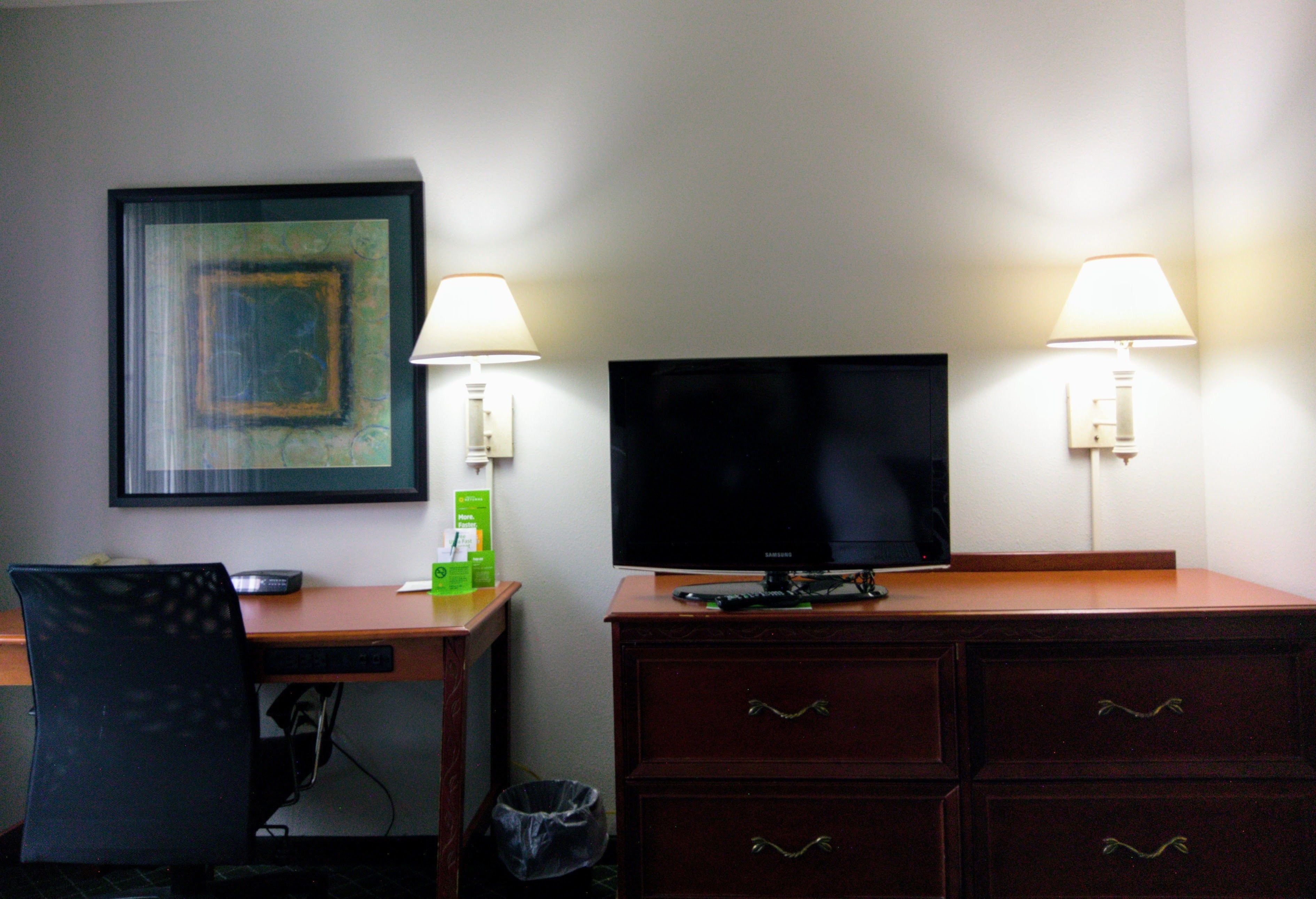 Quarto de hotel com escrivaninha, televisão, duas luminárias fixadas na parede, um quadro emoldurado e uma lixeira.