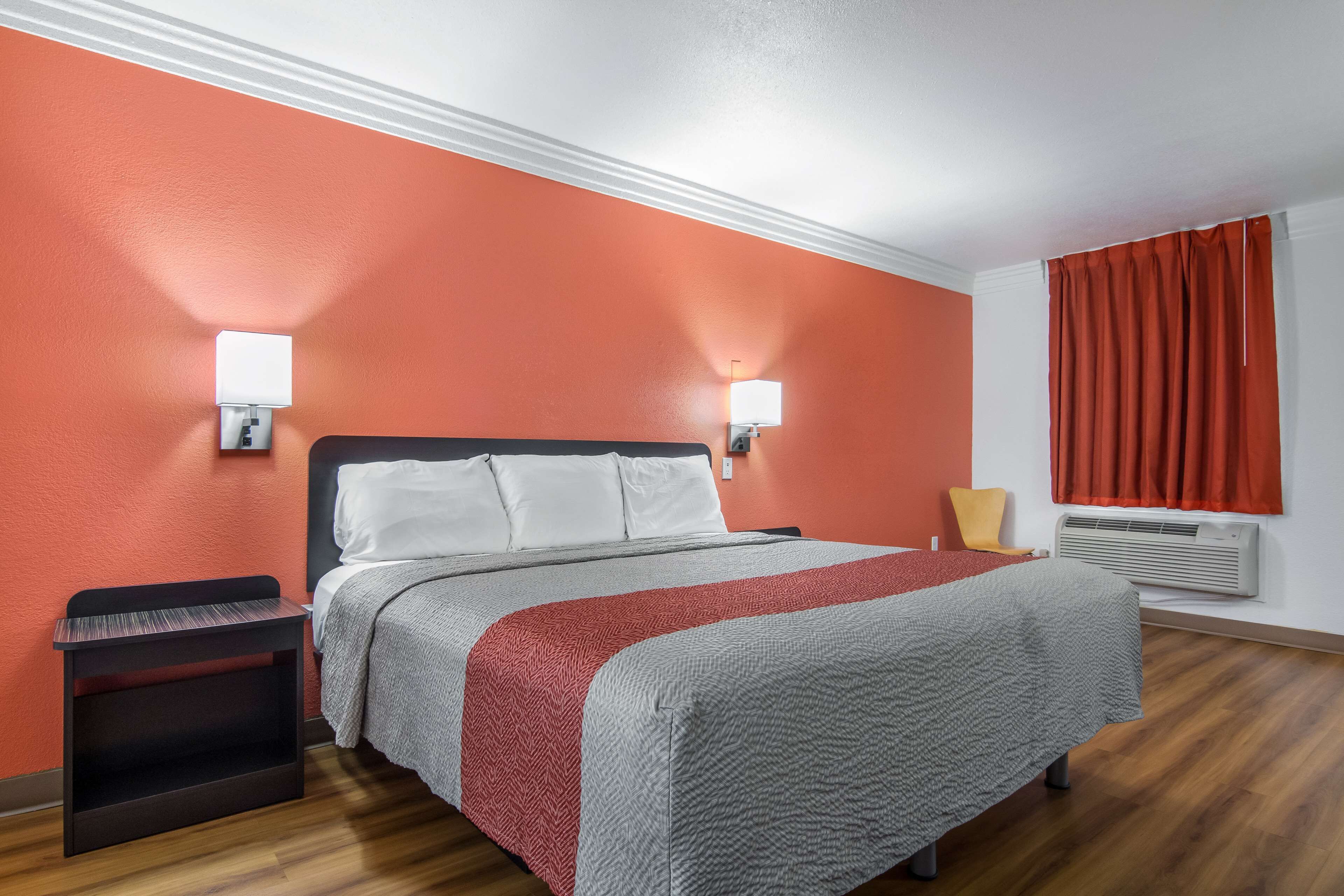 Quarto de hotel com cama, mesa de cabeceira, luminária e janela com cortinas.