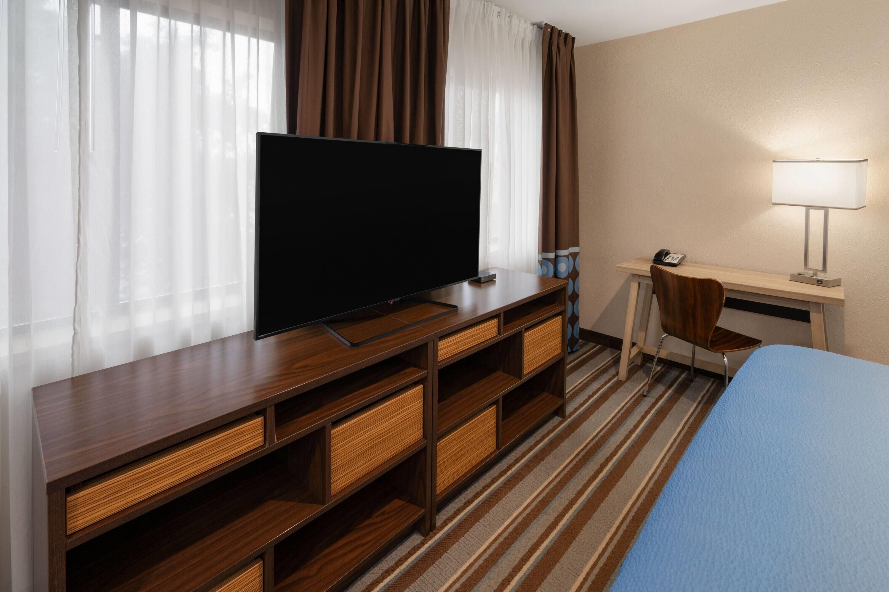 Quarto de hotel com televisão sobre um móvel de madeira, uma escrivaninha com cadeira e uma cama com colcha azul.