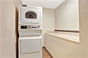 Máquina de lavar compacta fixada na parede, com visor digital e painel de controle, localizada no canto de um cômodo com piso de azulejos e um pequeno banco embutido.