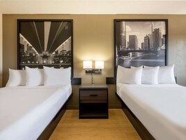 Quarto de hotel com duas camas, uma mesa de cabeceira com abajur, e duas quadros com paisagens urbanas na parede.